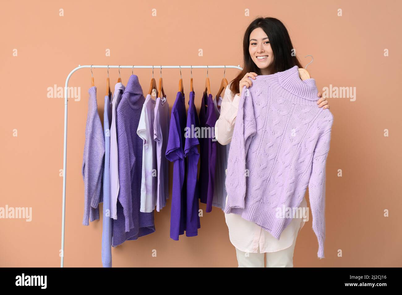 Young Asian woman and rack with clothes in purple shades on beige ...