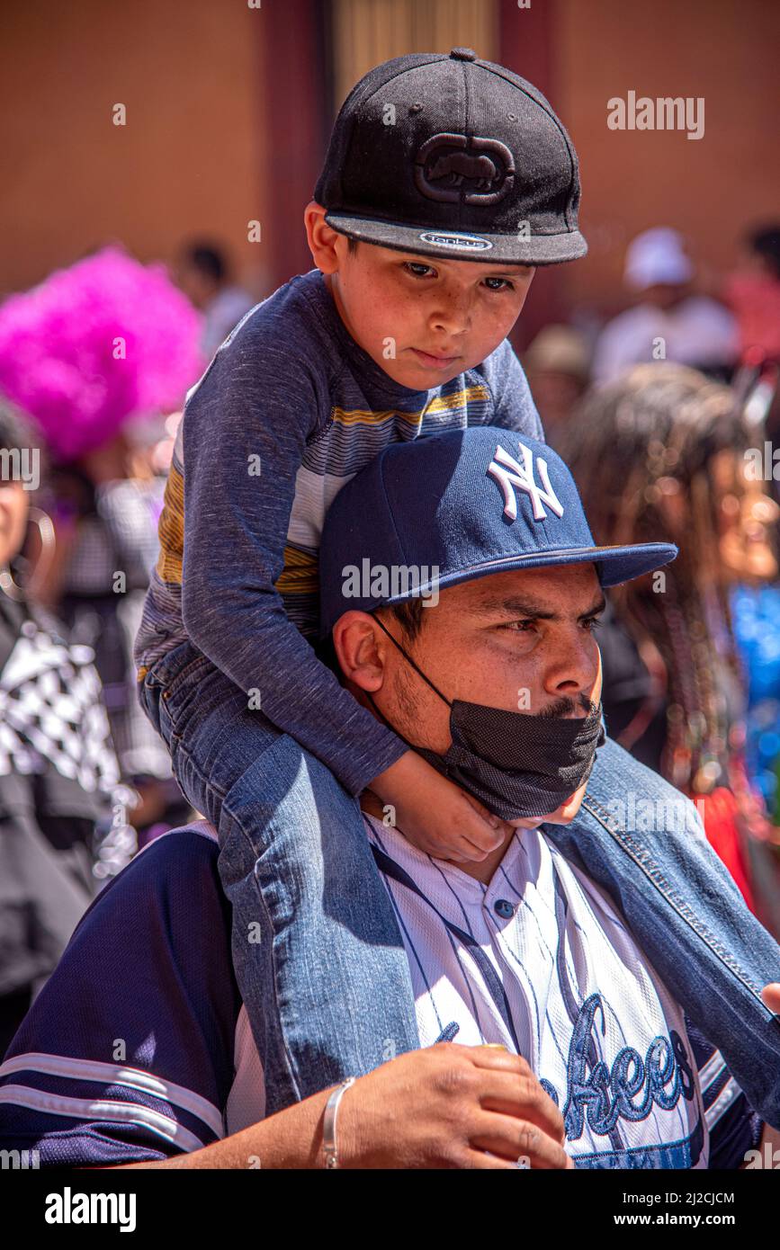 A Mexican father wearing a mask, carrying his son who is wearing a cap ...