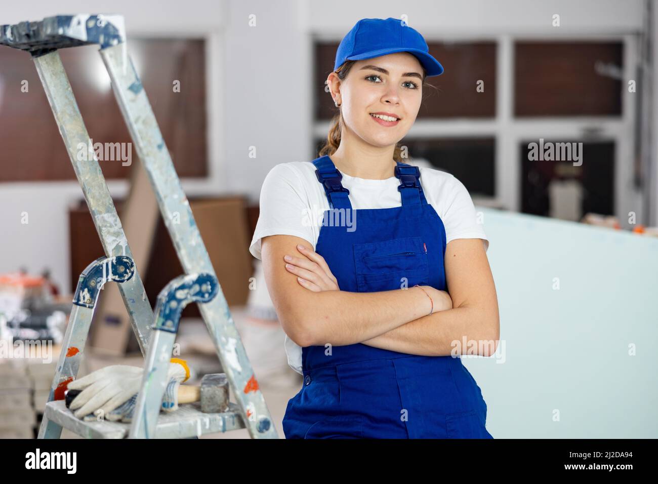 Portrait of positive builder woman in blue overalls next to stepladder ...
