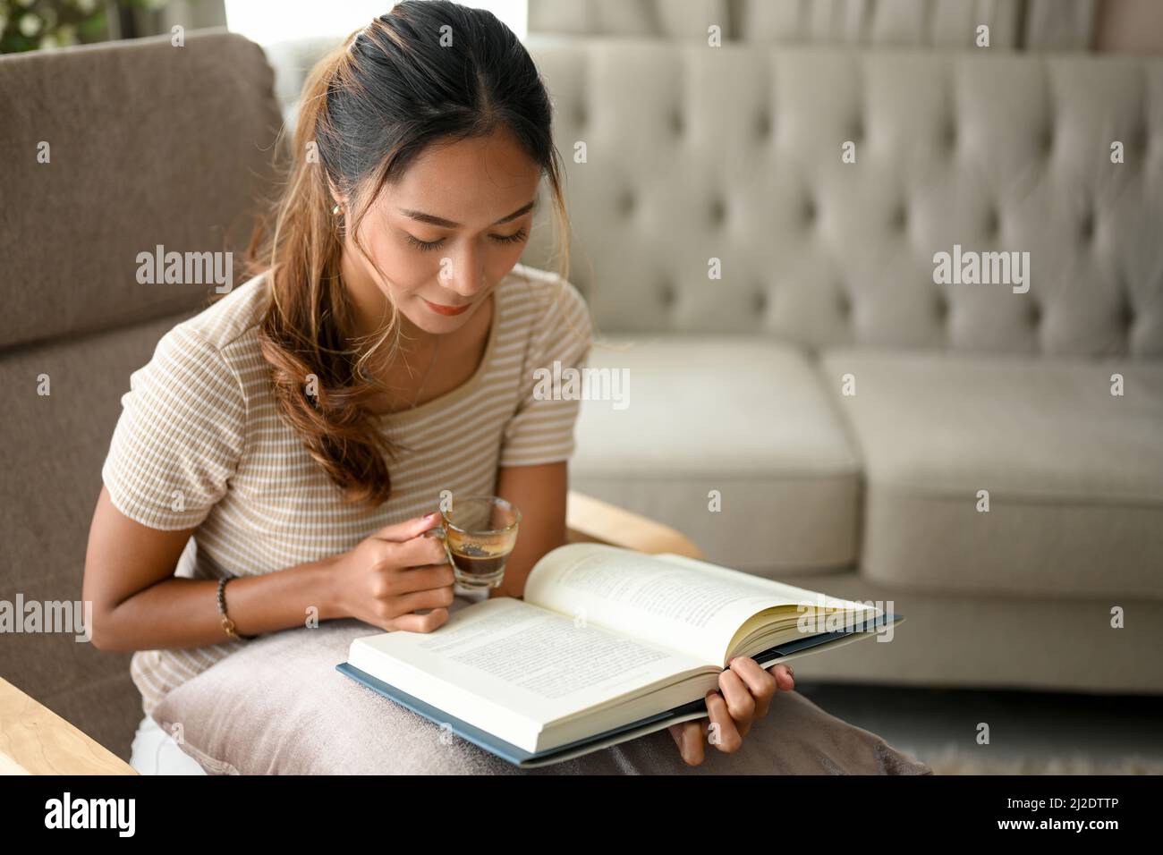 Beautiful Asian young woman in casual outfit reading a book in her modern living room. Hobby and ...