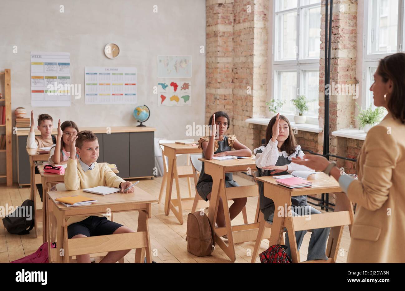 Diverse group of children raising hands in school classroom with young ...