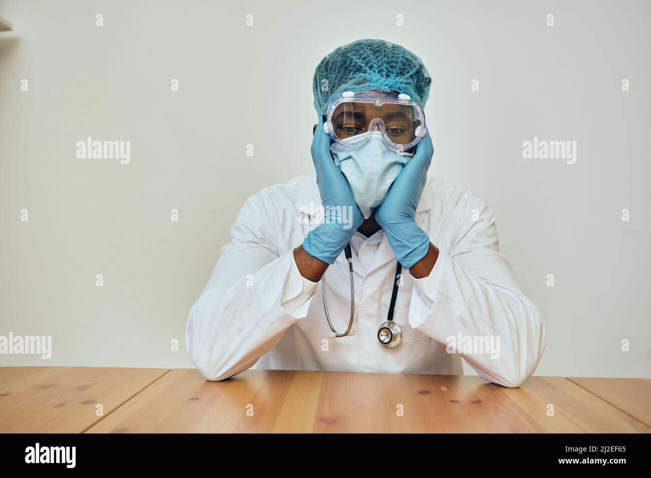 Male doctor wearing protection sitting at desk with hands clasped while ...
