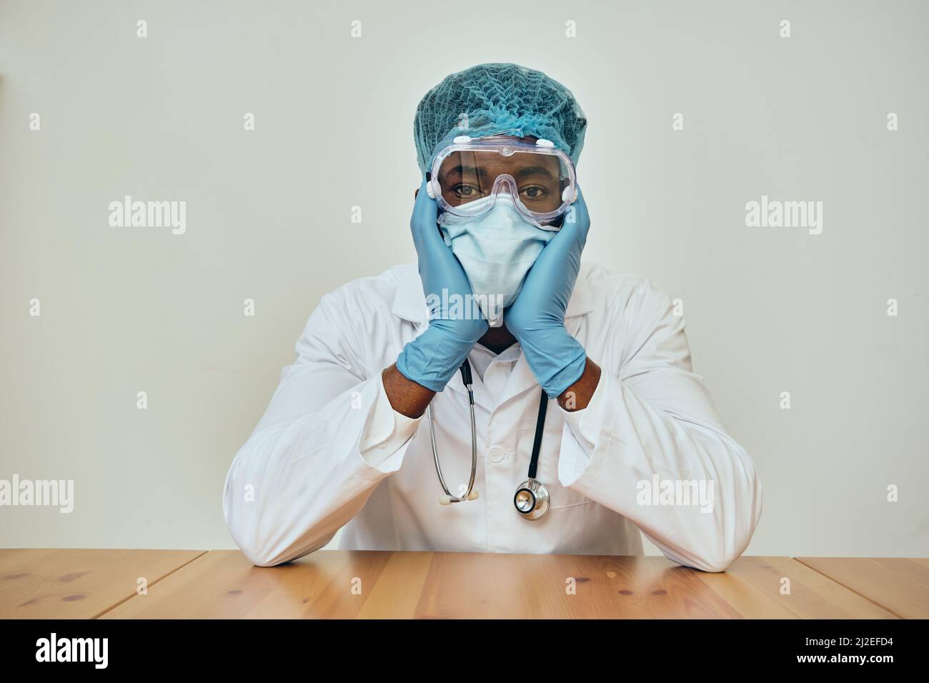 Male doctor wearing protection sitting at desk with hands clasped while ...