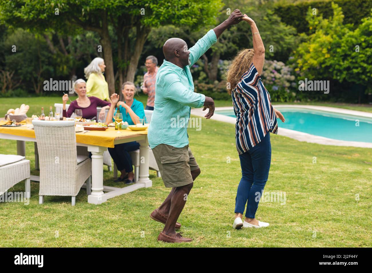 Multiracial senior couple dancing while friends at table in background ...