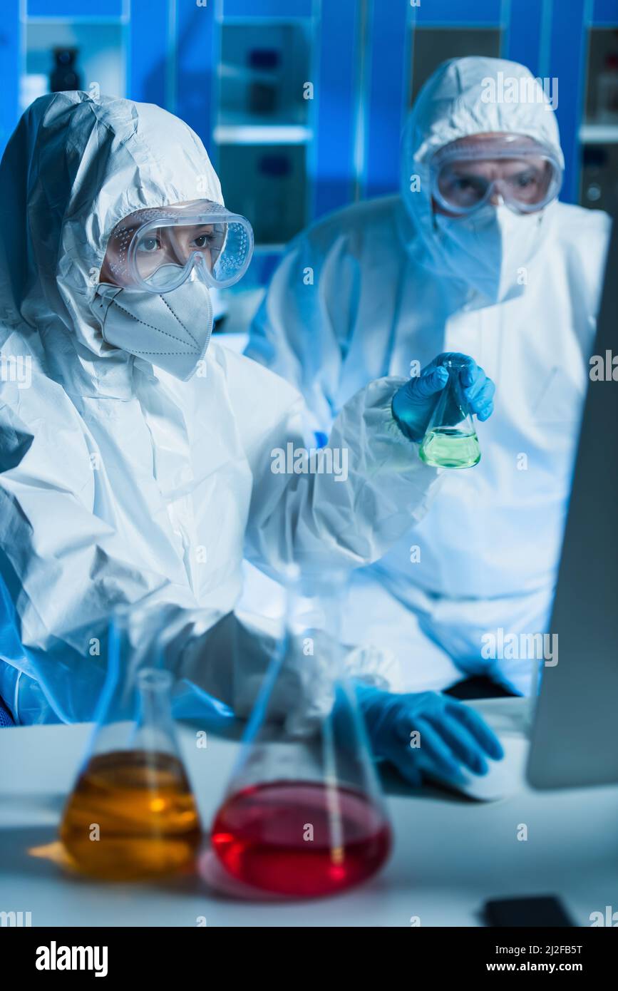 scientists in hazmat suits working in laboratory near blurred flasks ...