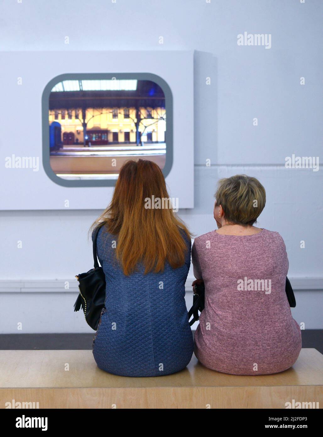 Two women sitting on a bench watching film on the screen at the ...