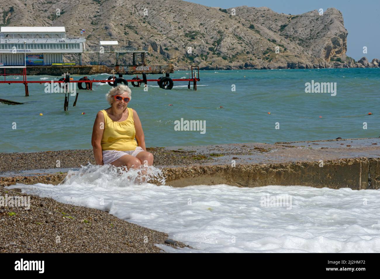 Plump smiling elderly woman in yellow blouse is sitting on beach pier ...