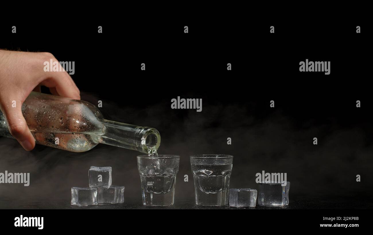 Bartender pouring up two shots of vodka with ice cubes from bottle into glasses on black ...