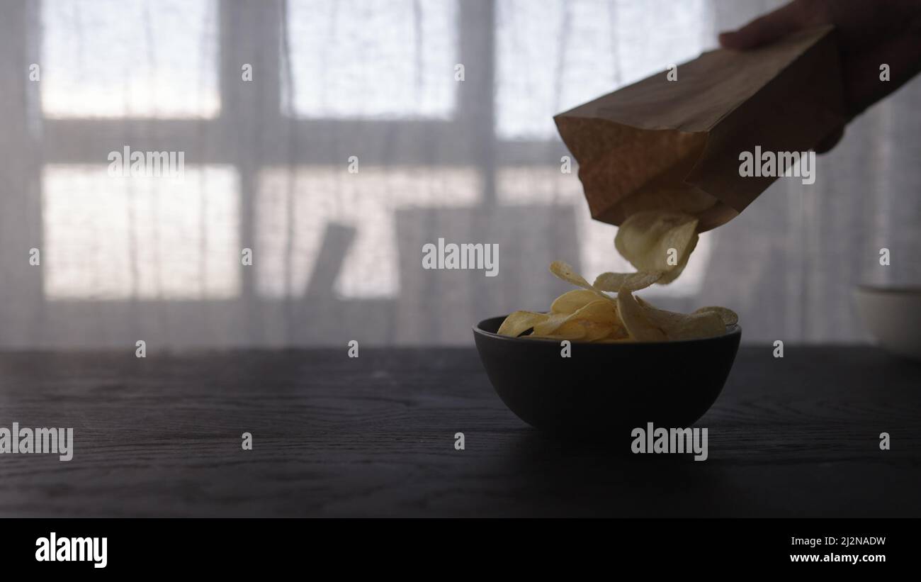 pour potato chips from paper bag into black bowl on oak table, wide ...