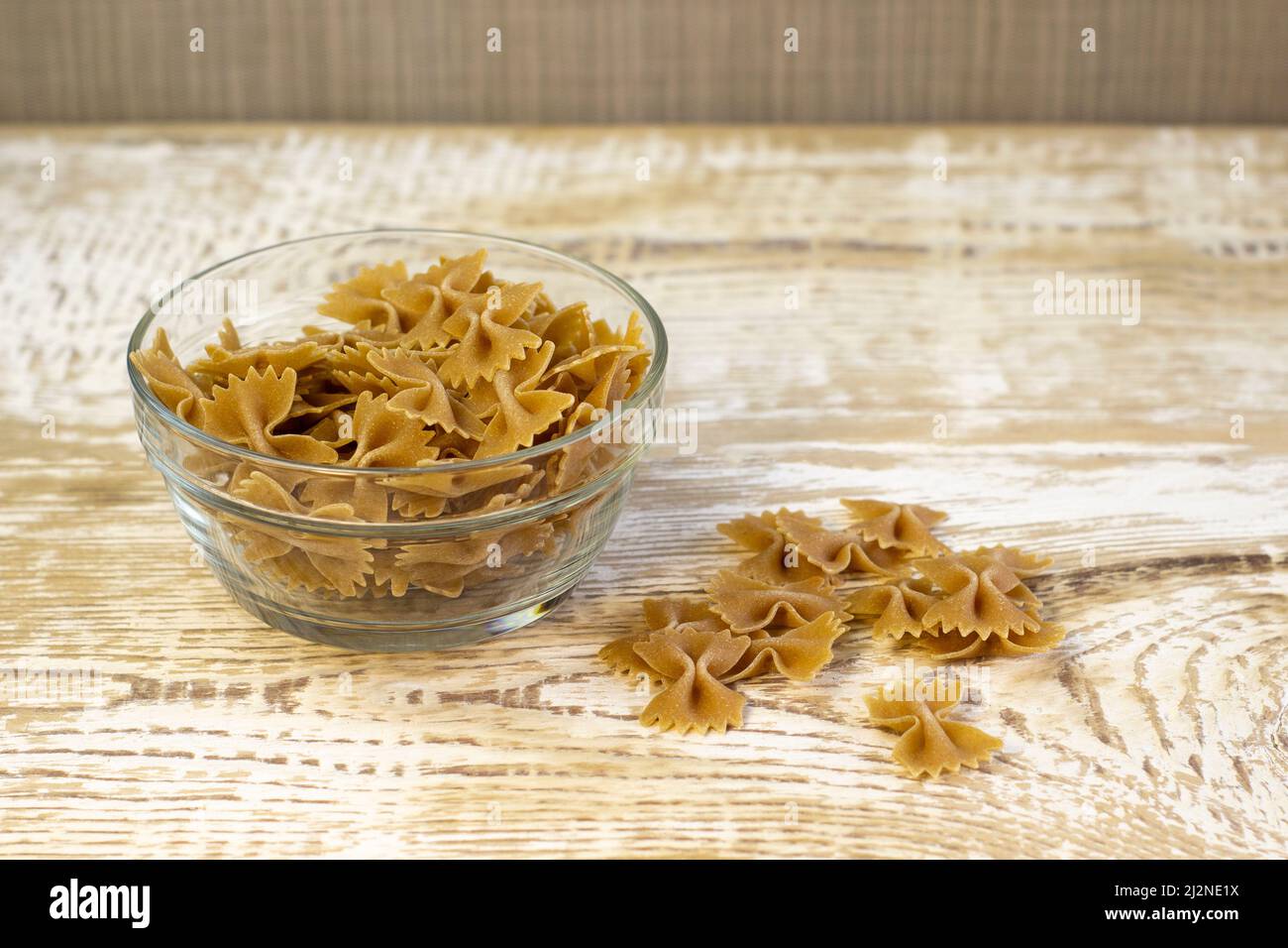 Whole grain dark pasta bows on a glass plate on a rough wooden light ...
