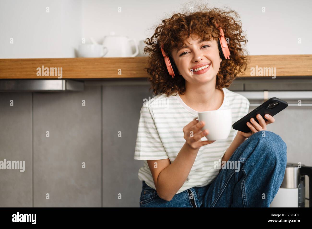 Smiling curly woman in headphones using cellphone while drinking coffee ...