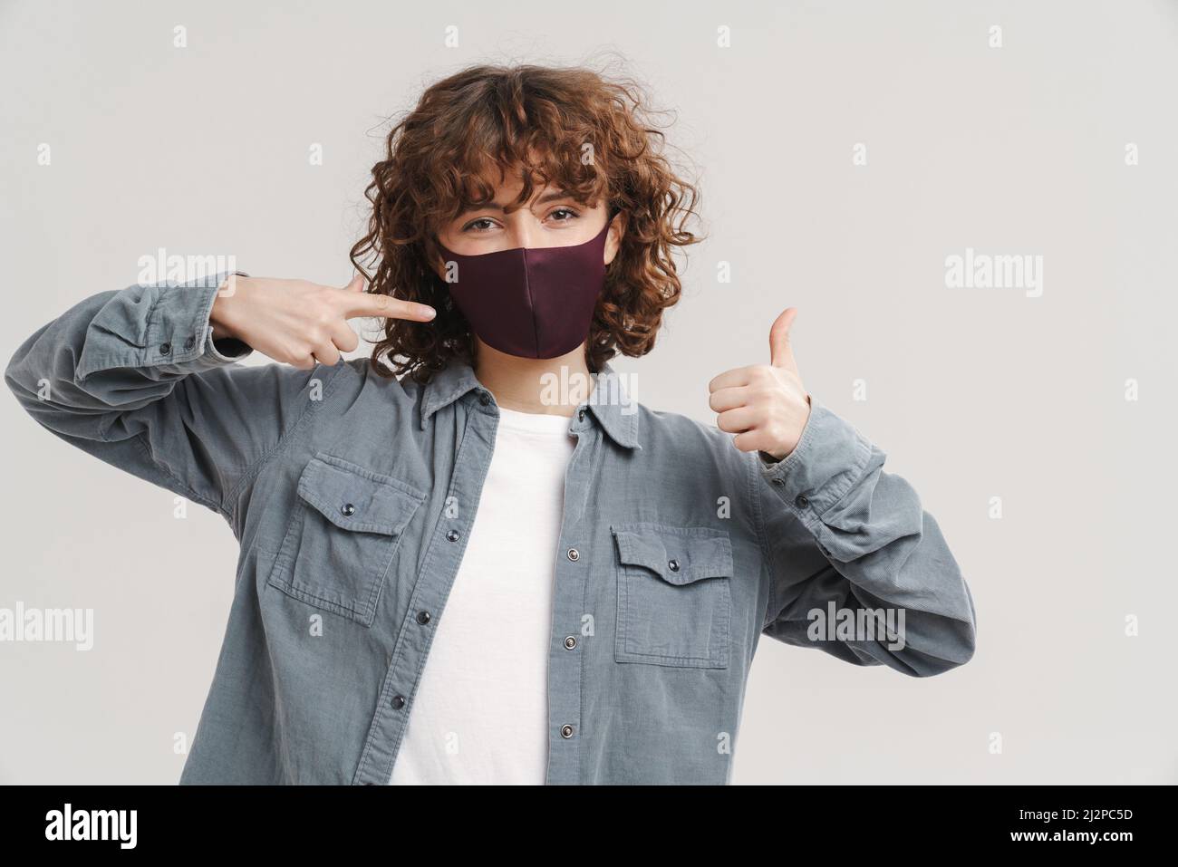 Pleased woman pointing finger at her face mask and showing thumb up ...