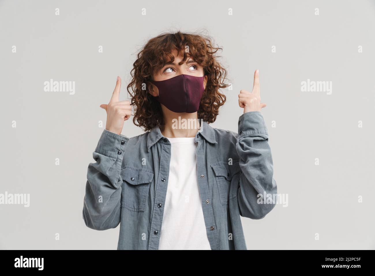 Young ginger woman in face mask pointing fingers upward isolated over ...