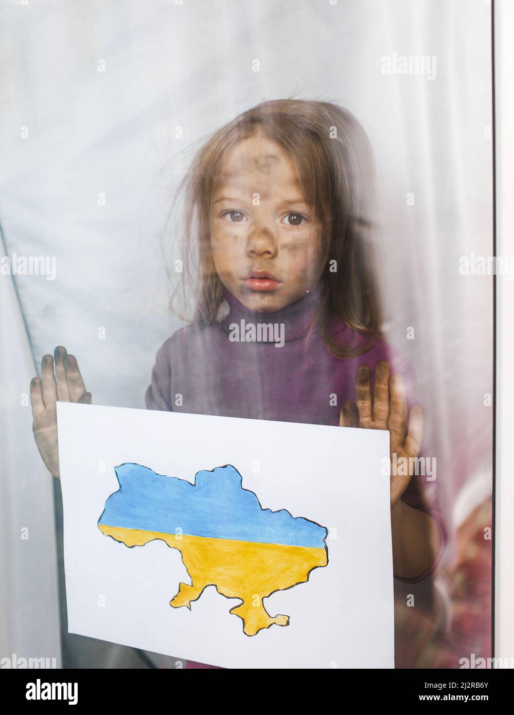 a little Ukrainian girl with sad eyes stands behind the glass, holding ...