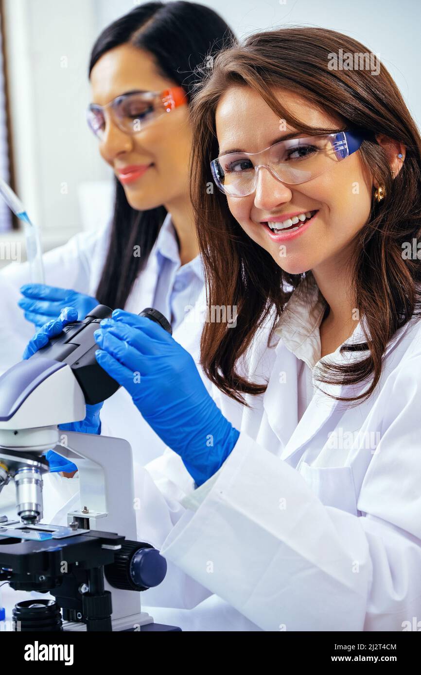 Young female scientist using microscope while her colleague working in background pharmaceutical ...