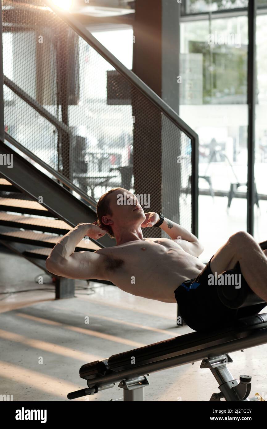Fit young man keeping hands behind head when doing decline sit-ups on ...