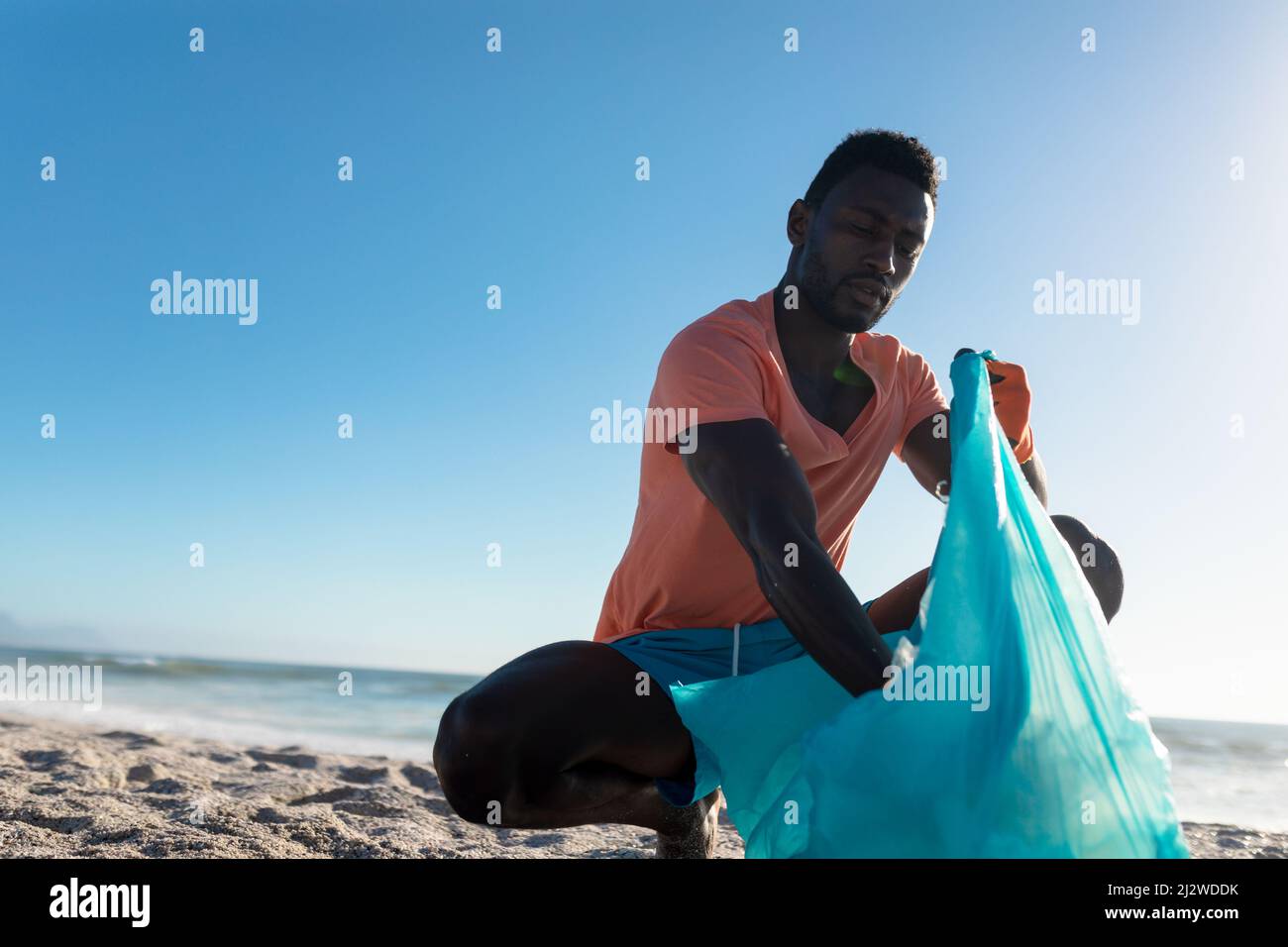 African american man crouching while collecting garbage in plastic bag ...