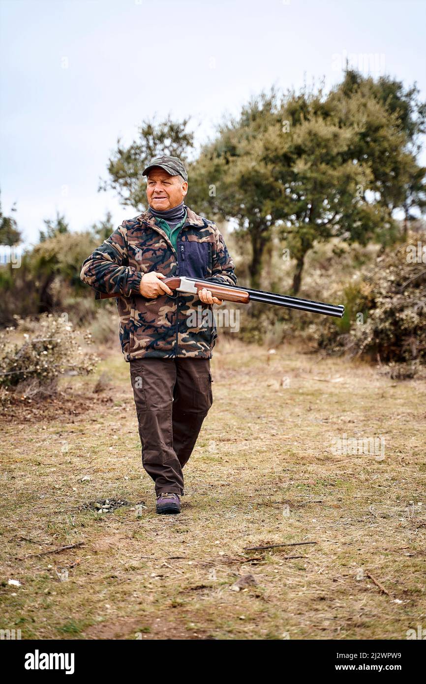 Full body aged male hunter in camouflage outerwear and cap carrying gun ...