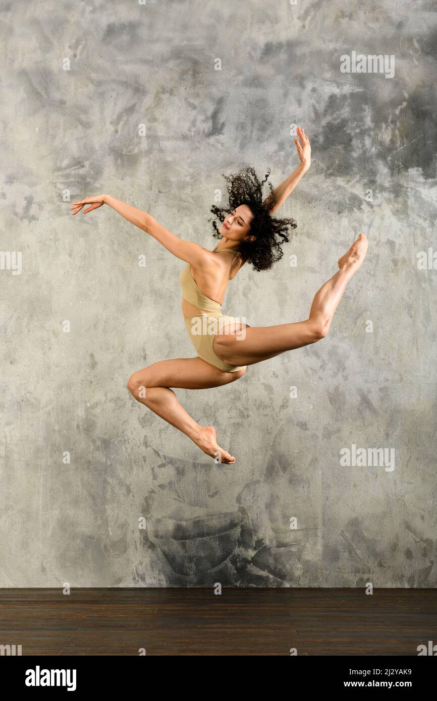 Full body side view of active barefoot female dancer in bodysuit performing jump with raised ...