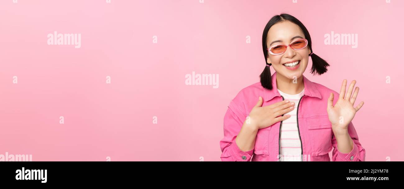 Portrait of friendly asian teen girl in sunglasses saying hello, waves ...