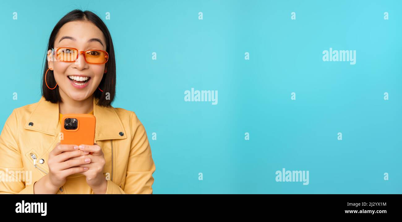Portrait of enthusiastic asian woman in sunglasses, using mobile phone ...