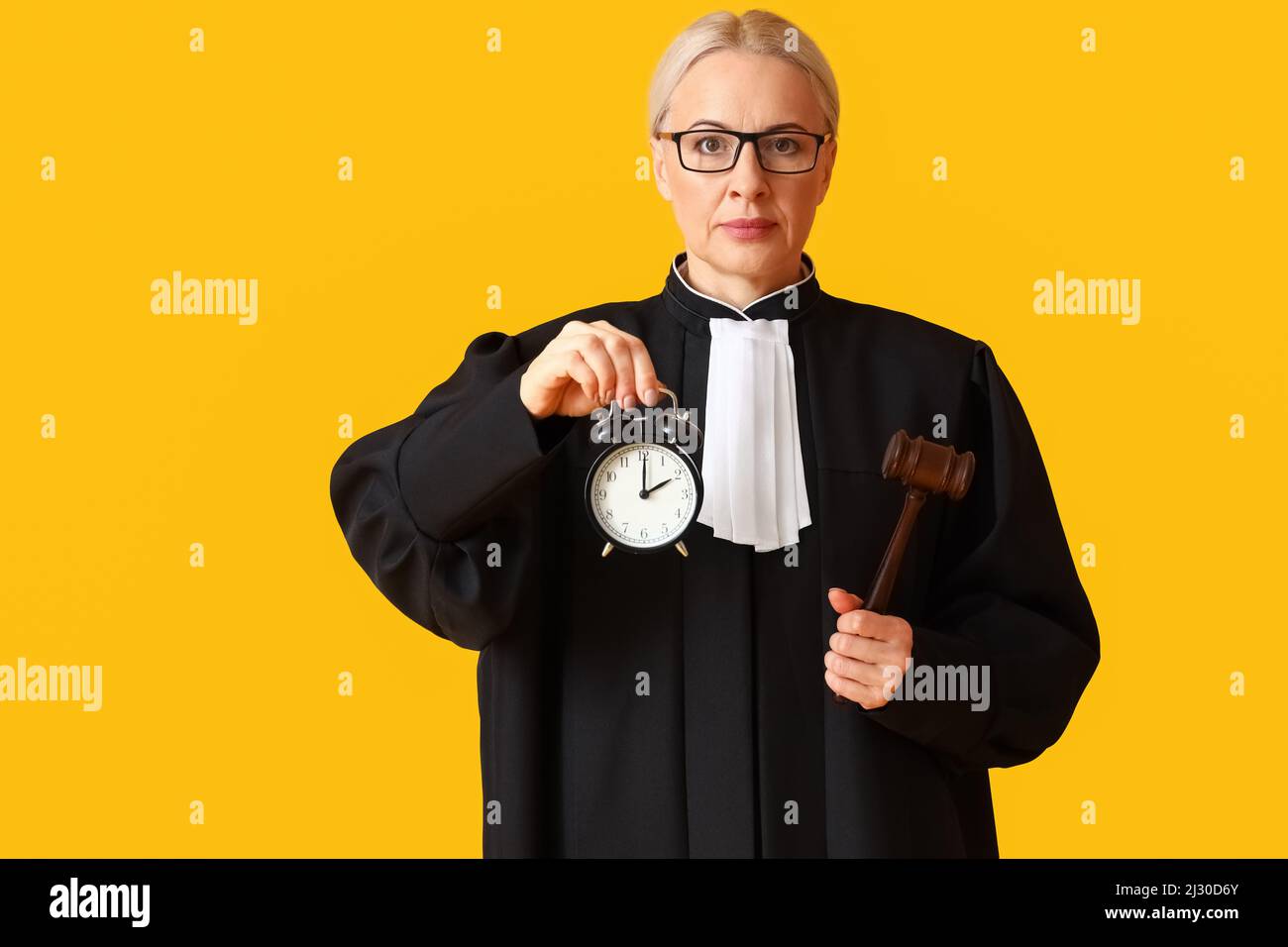 Mature female judge in robe with gavel and alarm clock on yellow ...