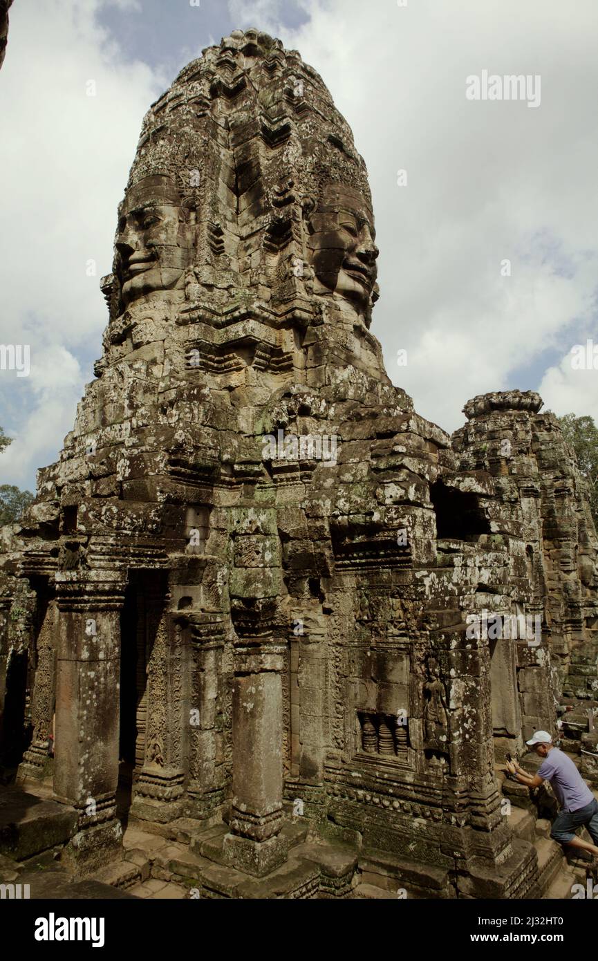 A tourist exploring Bayon temple below a stone tower carved with ...