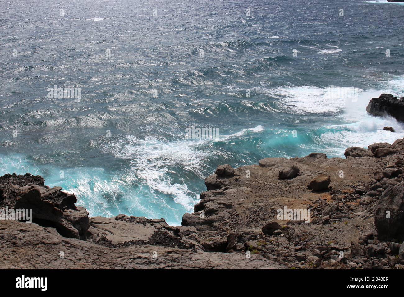 A volcanic rocky shoreline contrasted against varying shades of blue of ...