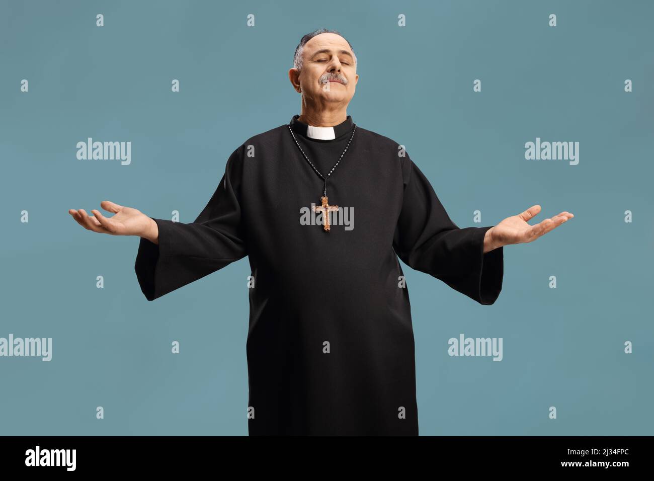 Mature priest with closed eyes spreading arms isolated on blue ...