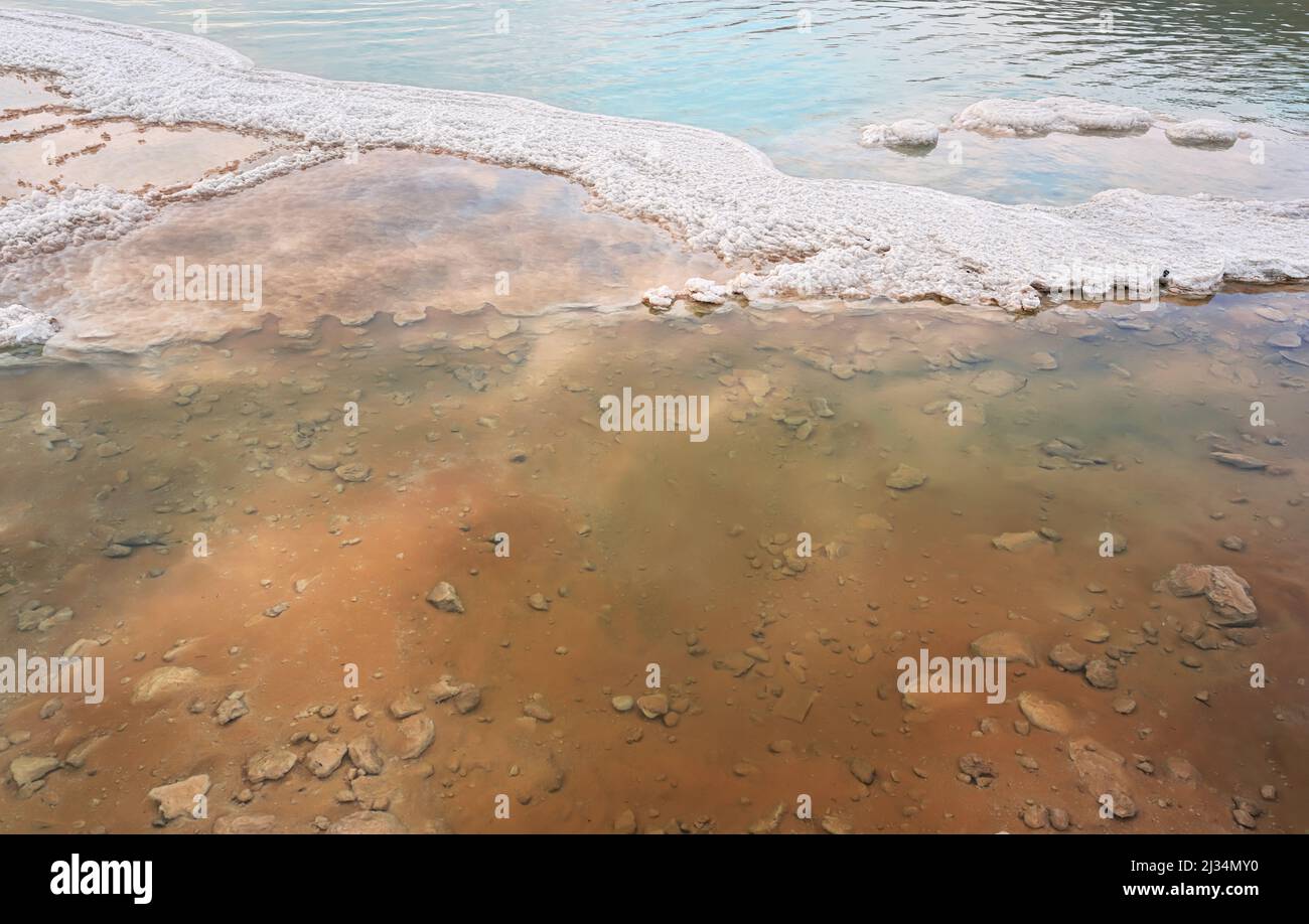 Sand covered with crystalline salt on shore of Dead Sea, clear calm water near - typical scenery ...