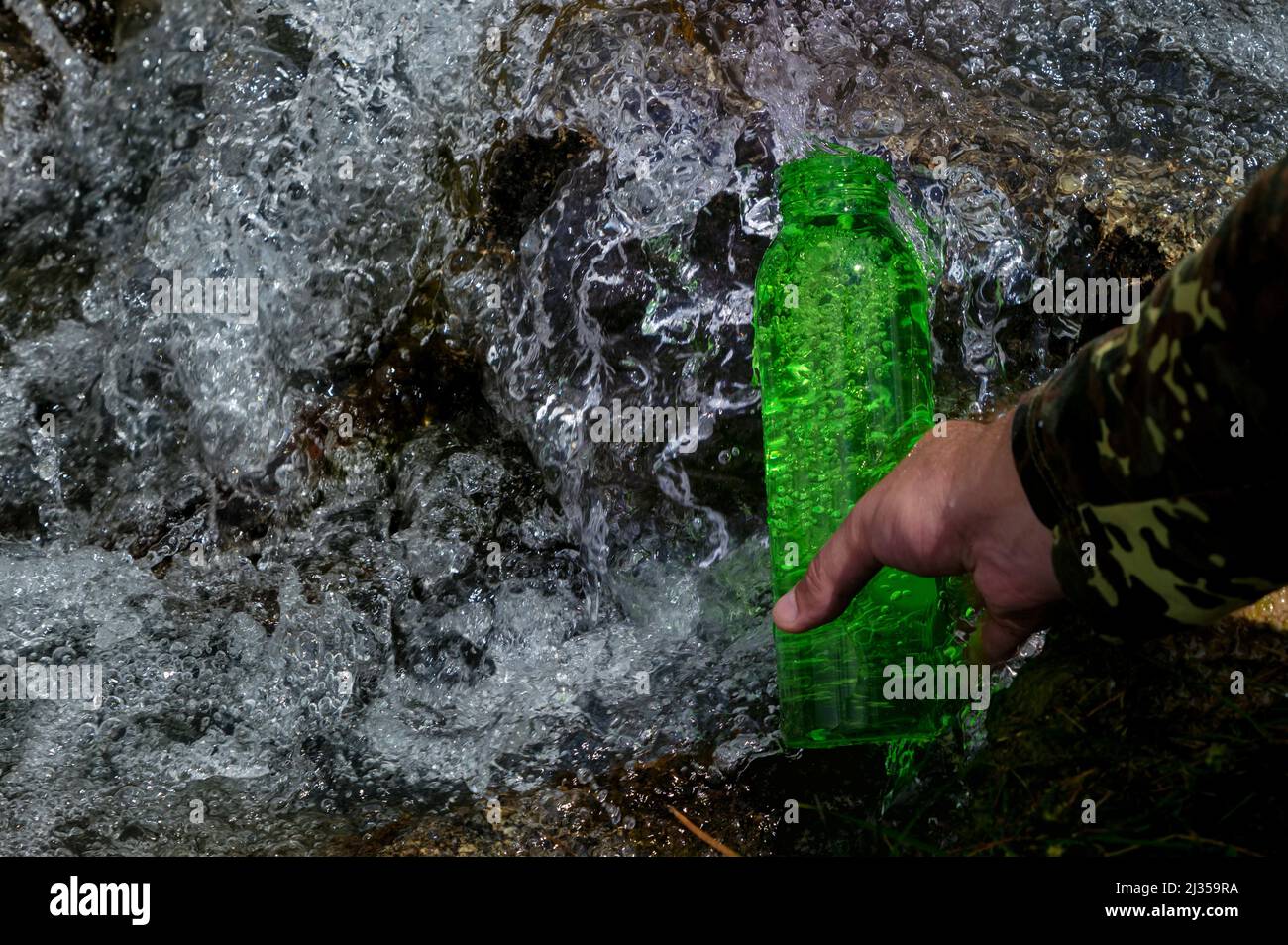 Hand of a traveller holds bottle collecting water from mountain stream ...