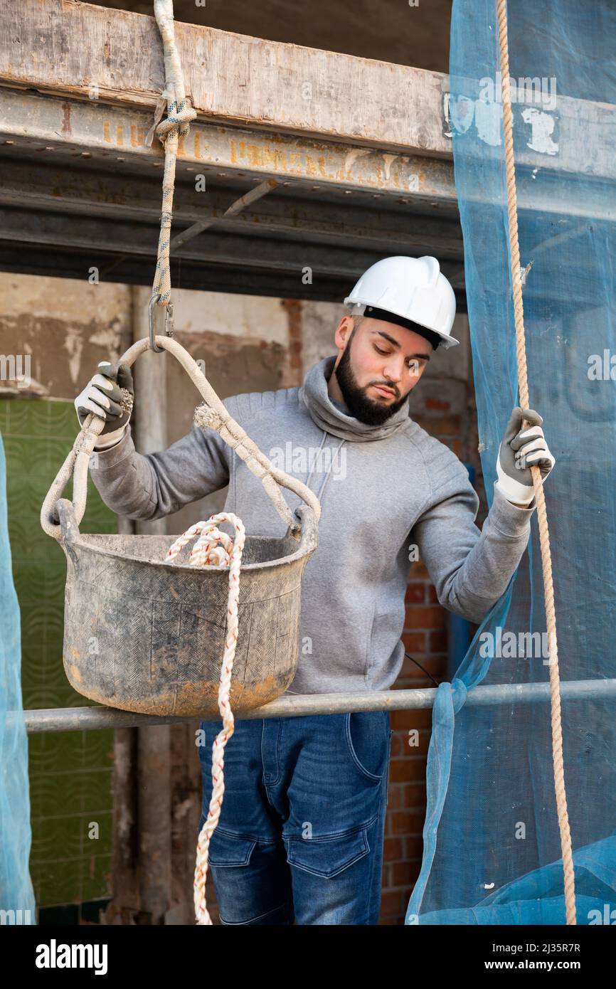 Worker lowering down bucket with construction mortar Stock Photo - Alamy