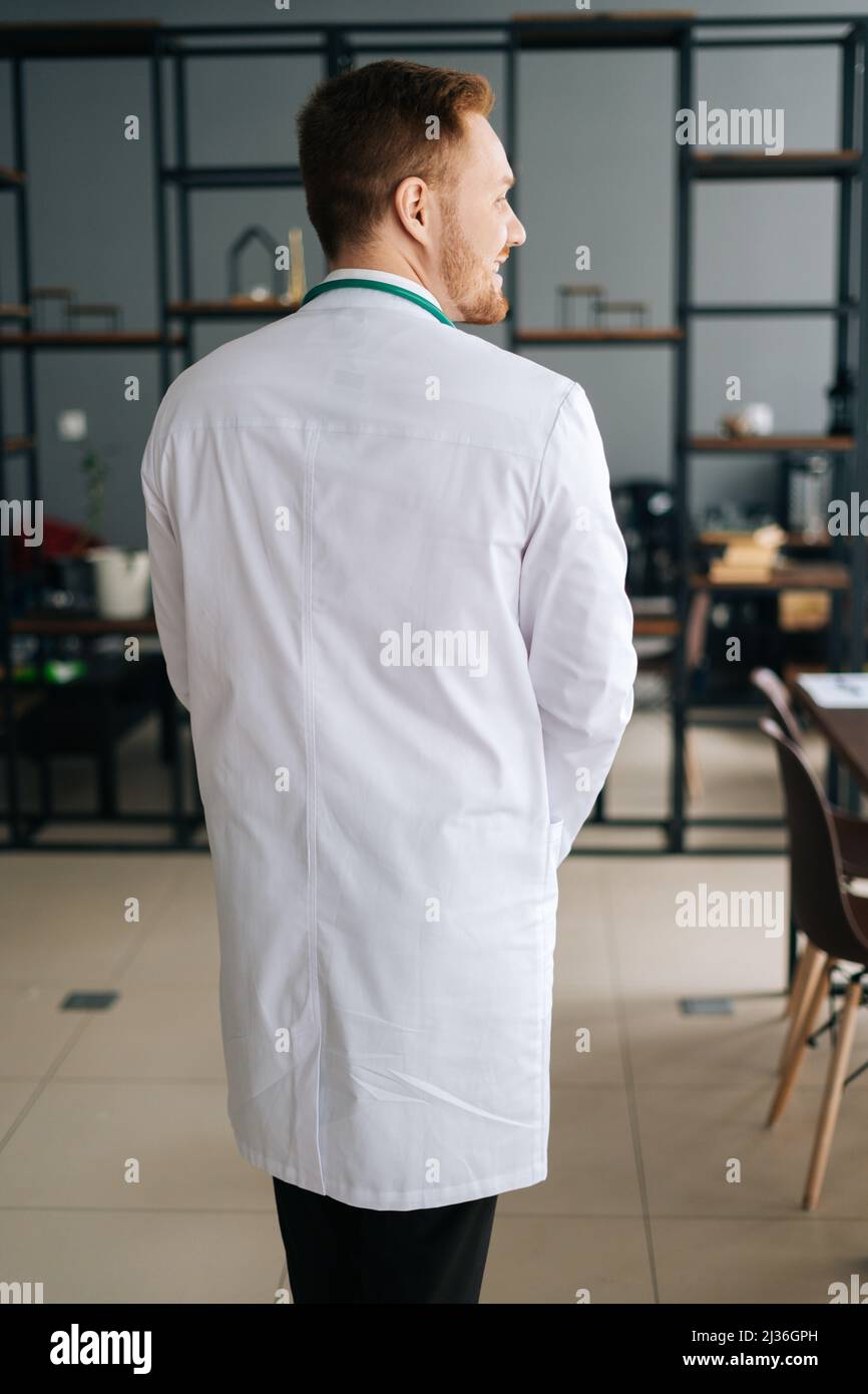 Rear view of smilng young male doctor in white medical uniform standing ...