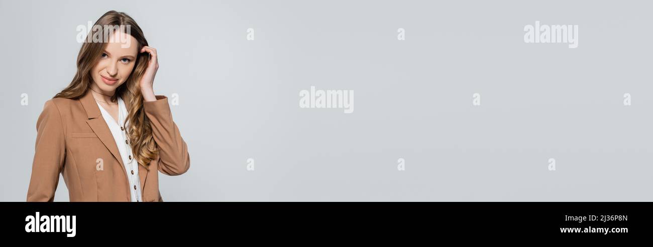 positive woman fixing long hair while looking at camera isolated on ...