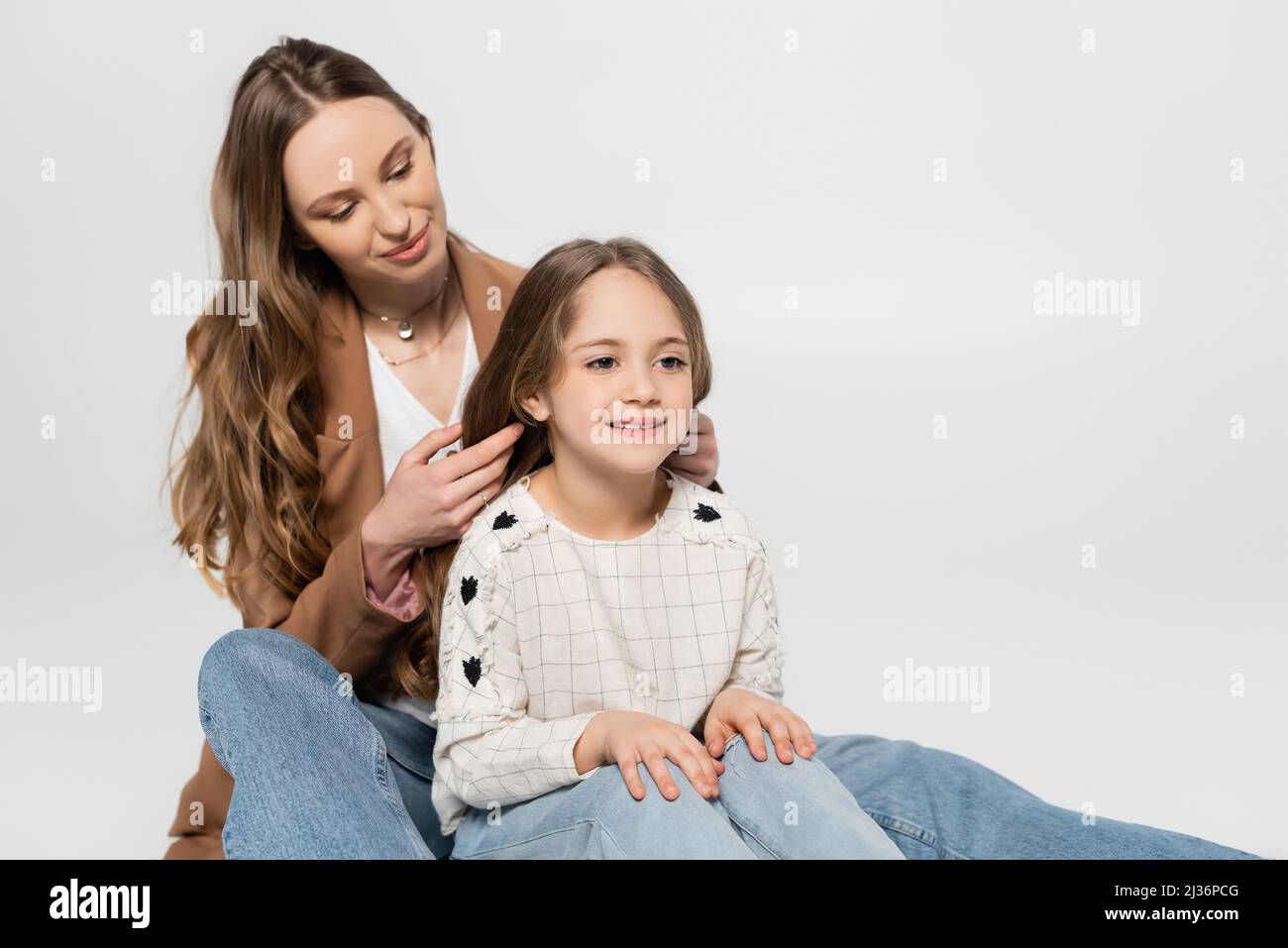 woman braiding long hair of cheerful daughter isolated on grey Stock ...