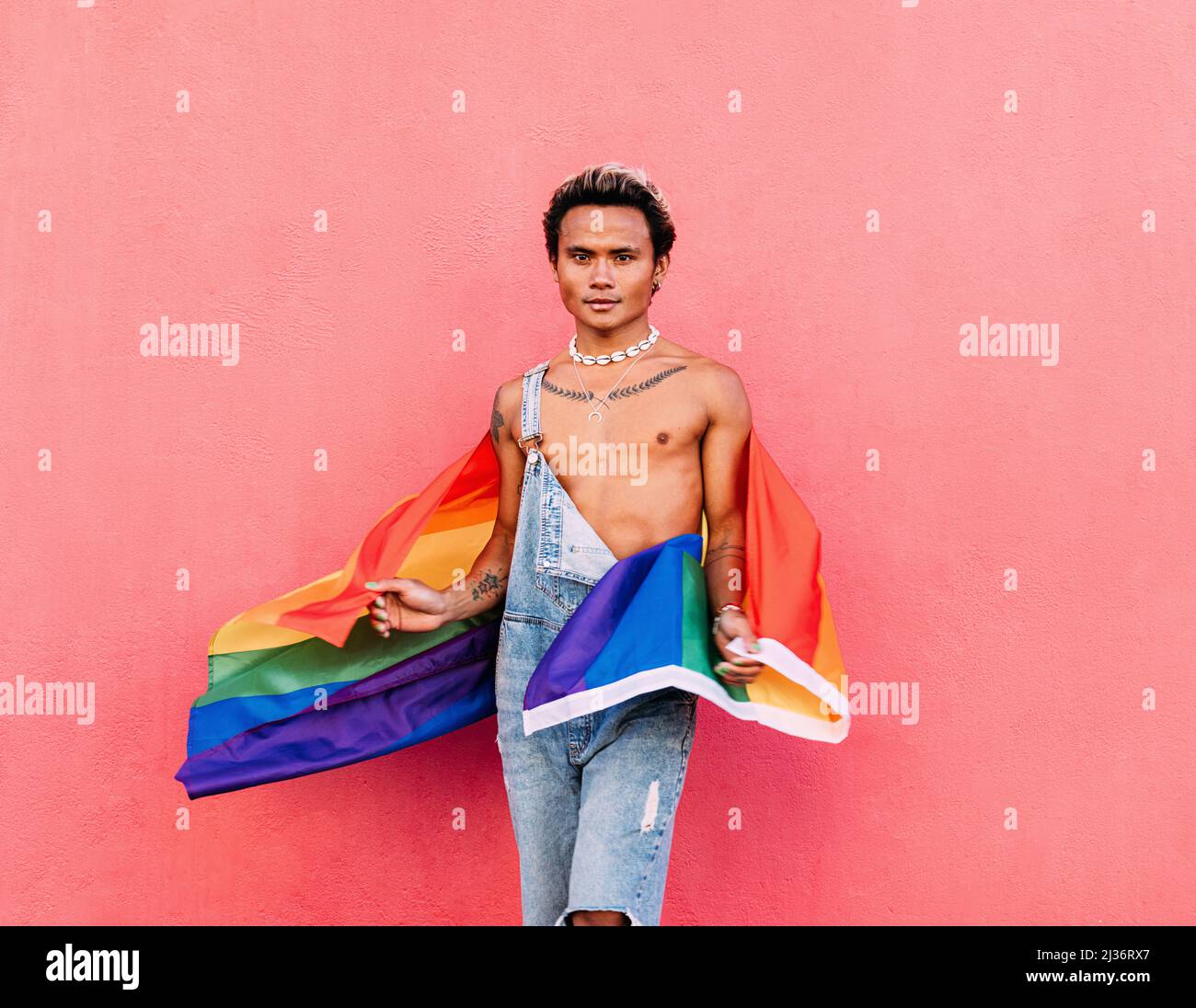 Stylish guy in denim overalls holding LGBT flag and looking at camera ...