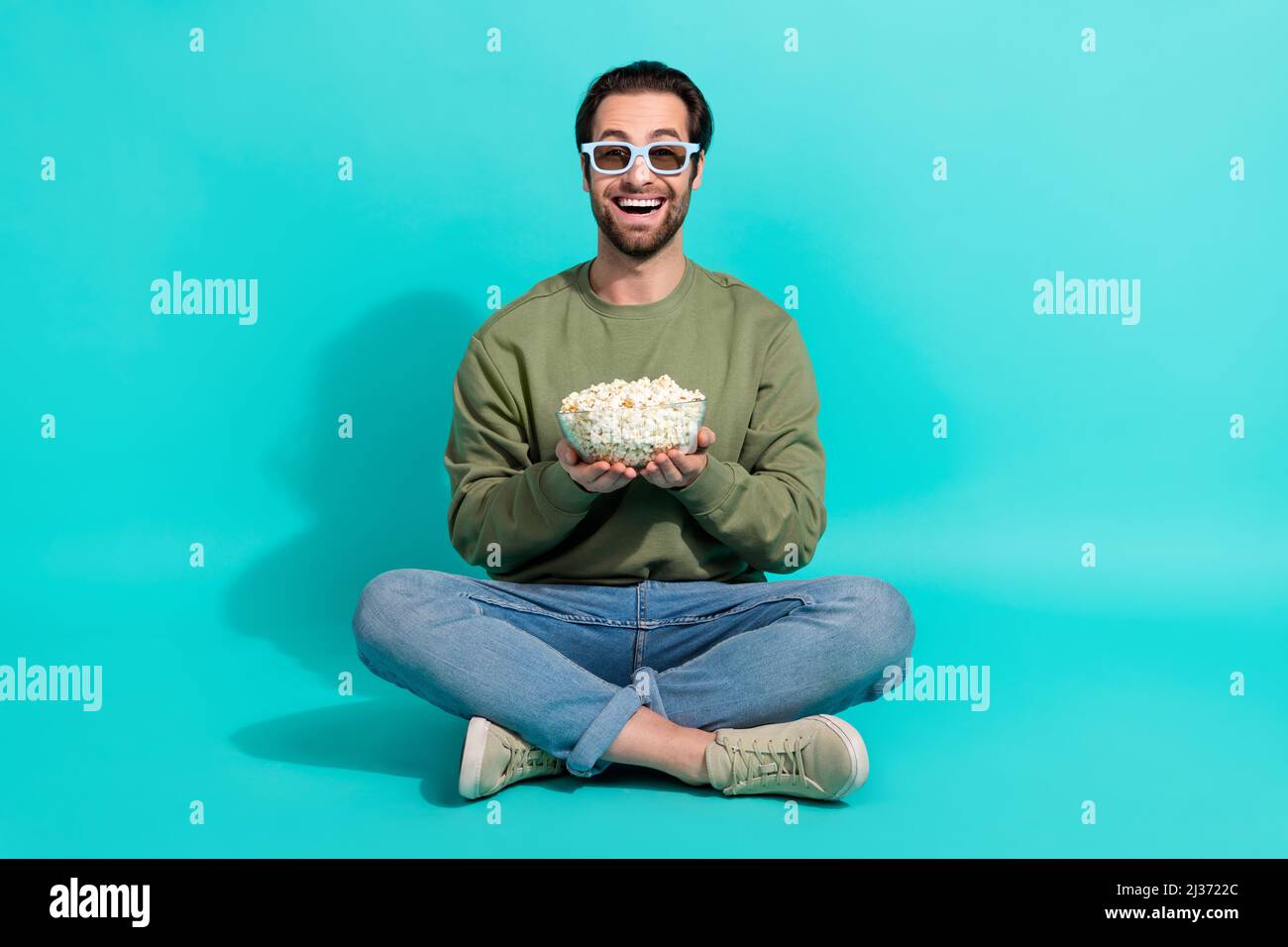 Full size photo of young cheerful man sit floor eat pop corn watch film ...