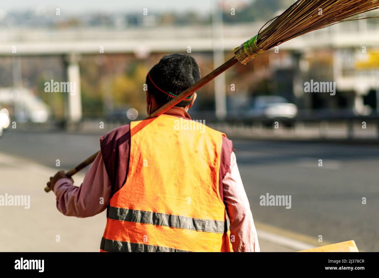 Back view of a city janitor in an orange uniform with a broom on his ...