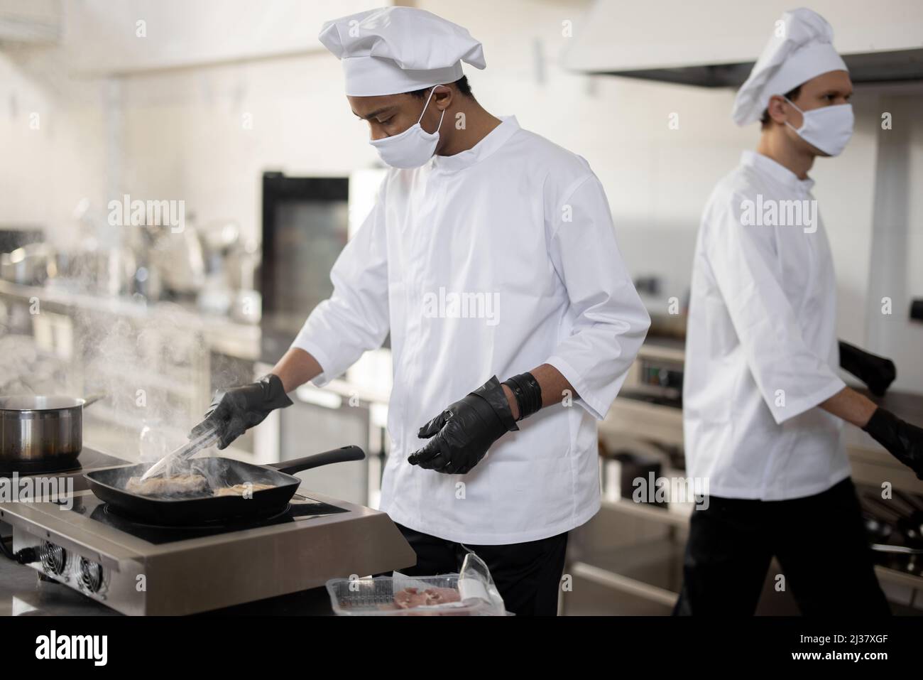Two chefs in uniform and face masks cooking together in professional ...