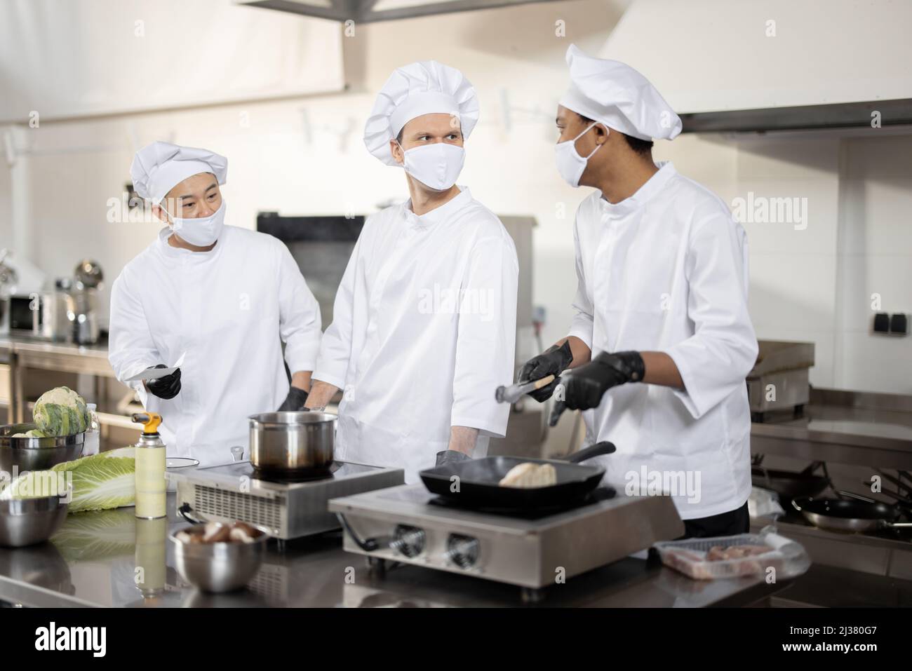 Multiracial team of cooks in uniform and face masks cooking meals for a ...