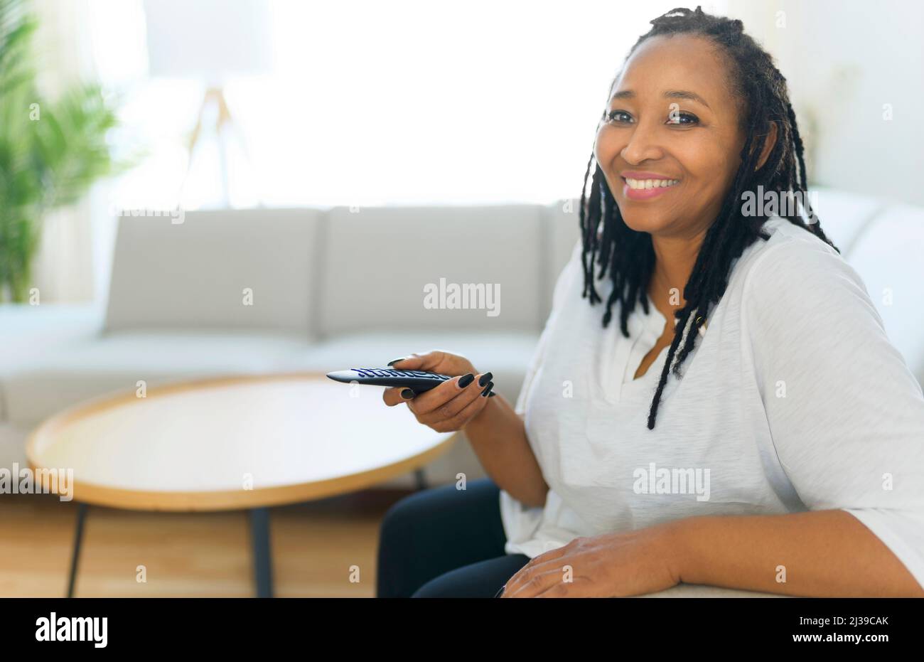 Portrait of an african woman sit on the sofa and holding remote control ...