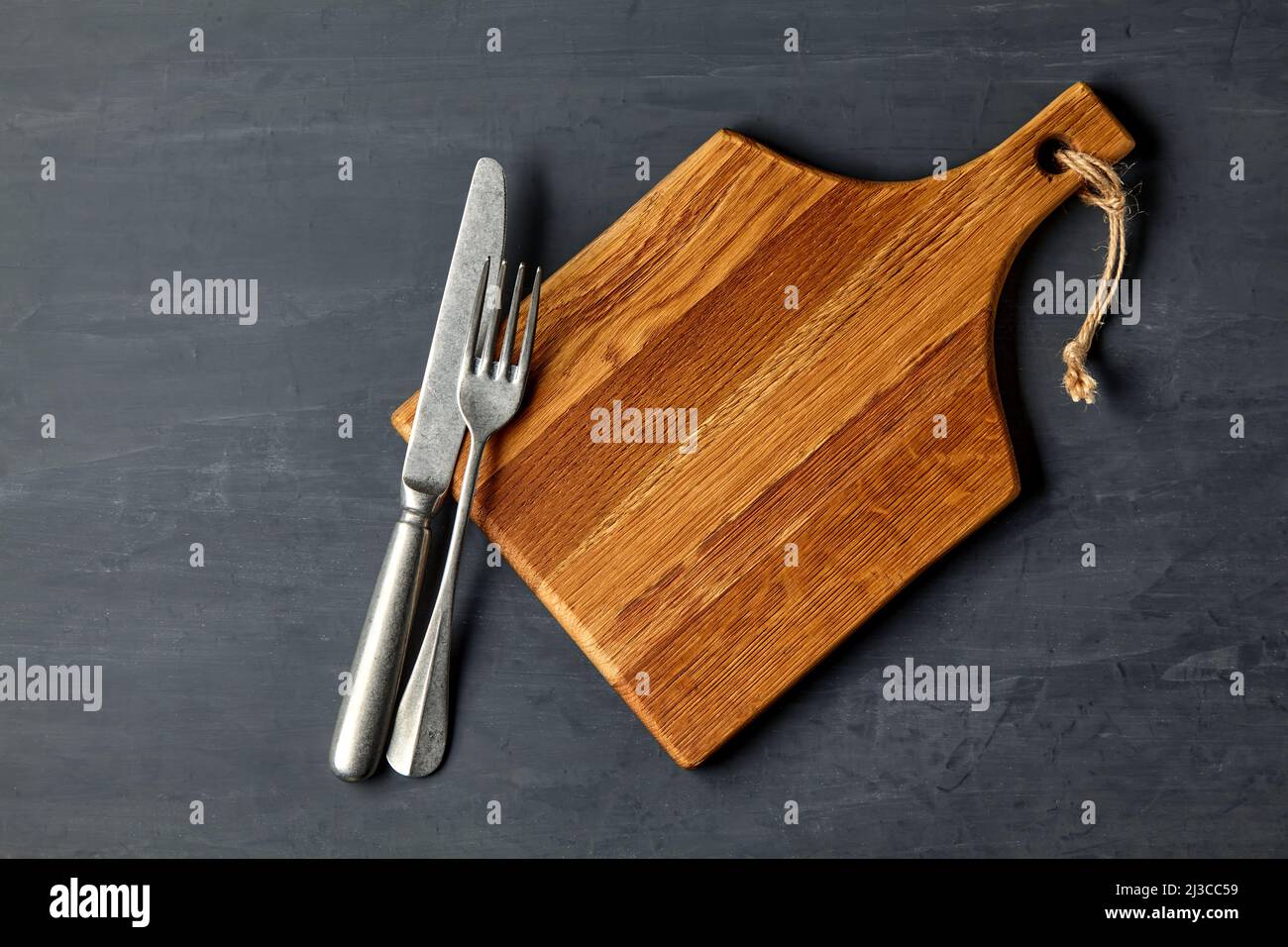 Brown wooden cutting board with cutlery on a dark concrete table ...