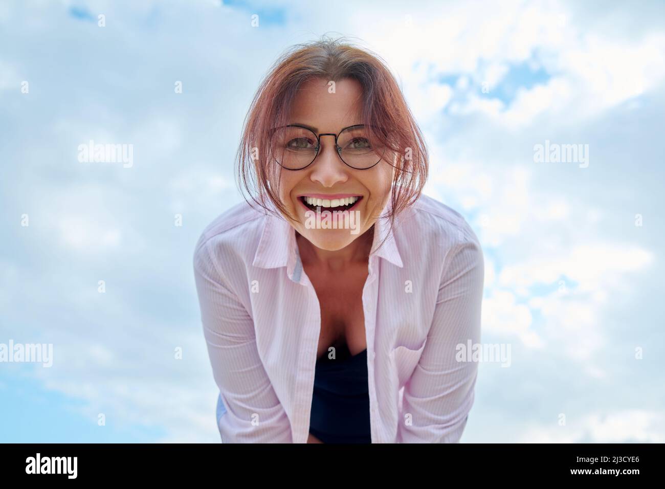 Happy middle age woman looking down at camera, blue sky in clouds ...