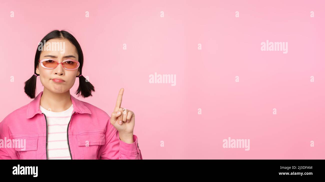 Close up of stylish korean girl, looking perplexed, pointing fingers up ...