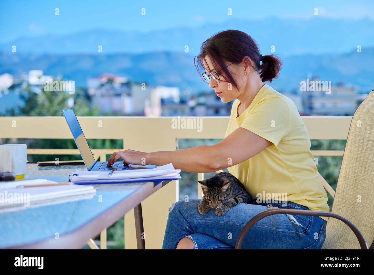 Middle-aged woman working in home office on terrace with pet cat in her ...