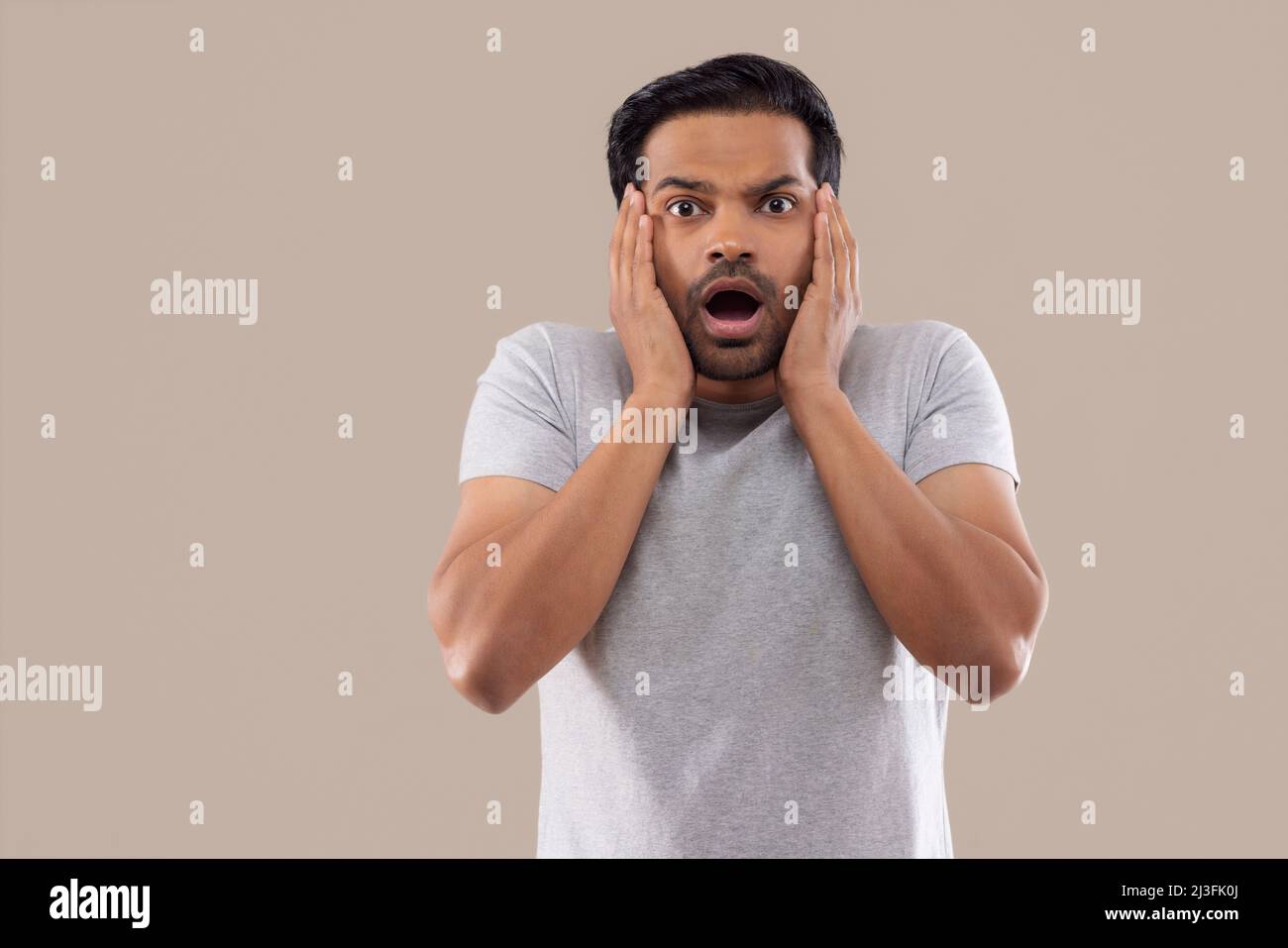 Portrait of a surprised young man with mouth opened and hands on cheek ...