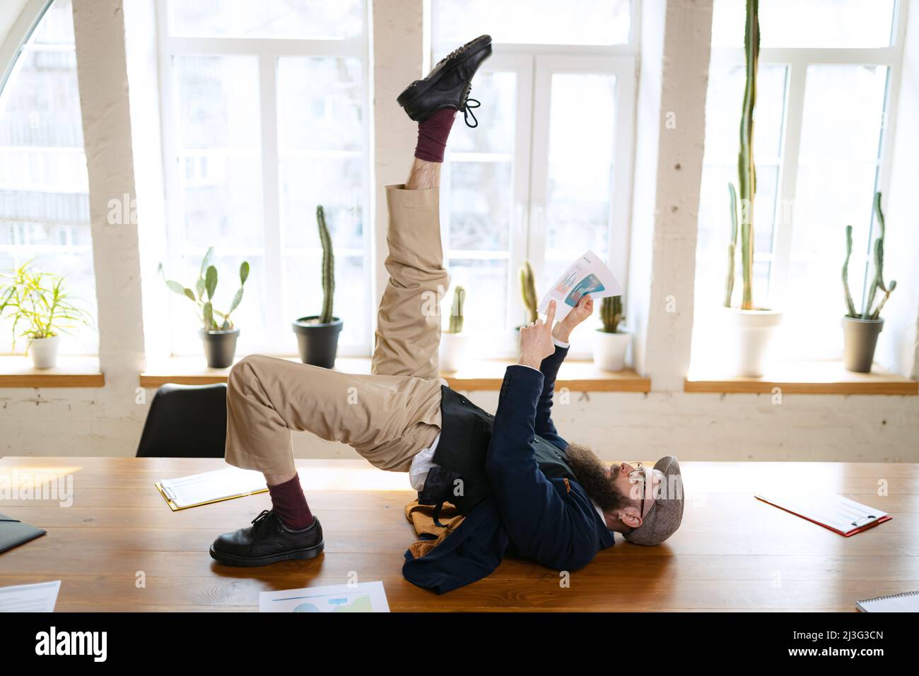 Young bearded man, office clerk having fun, doing yoga on wooden table ...