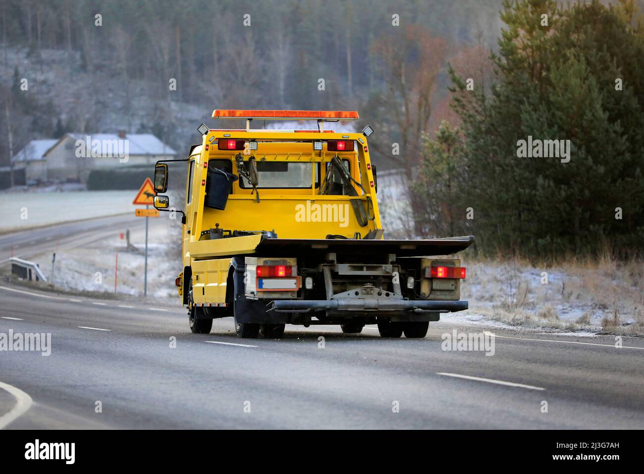 Yellow flatbed tow truck at speed on highway on a day of winter, rear ...