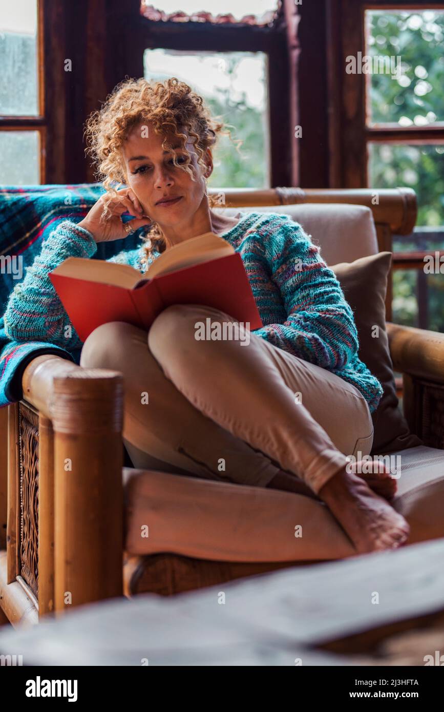 Relaxed woman alone at home sitting on a chair and reading a red book ...