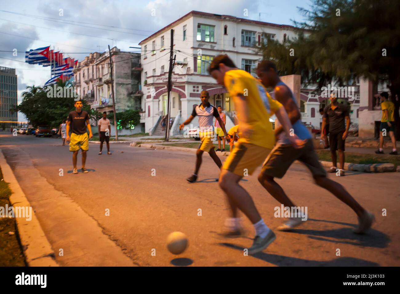 At dusk, young men play a game of soccer (futbol, football) on a street ...